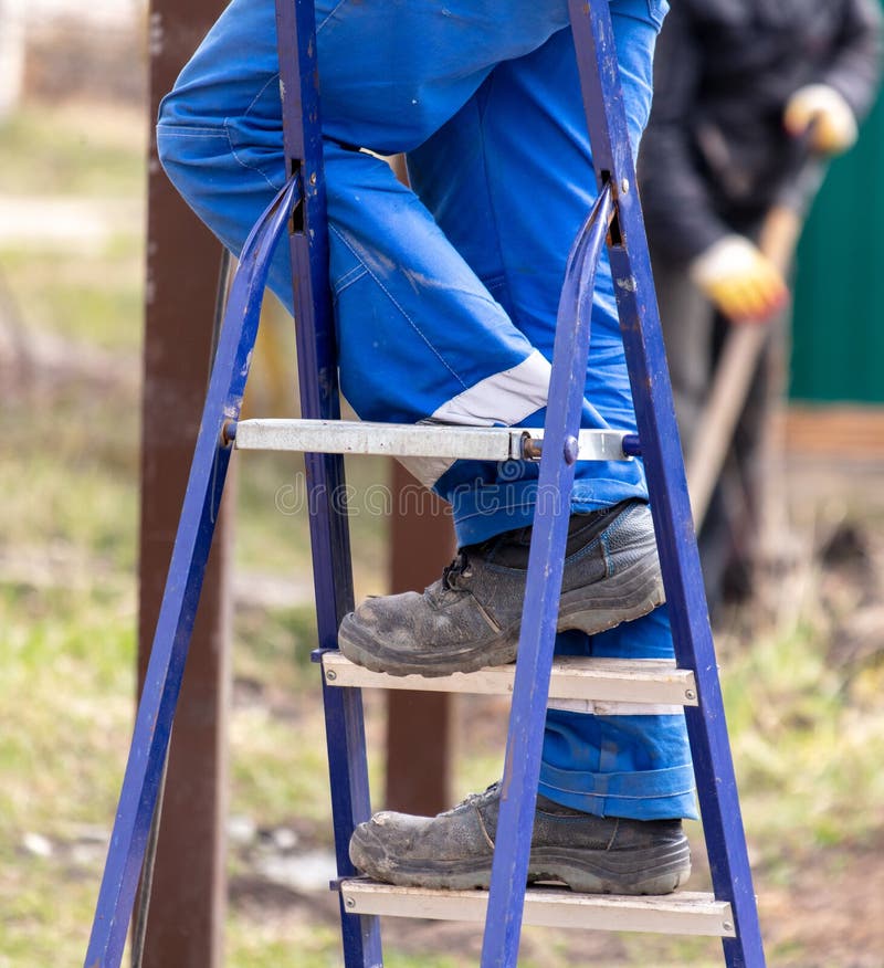 Worker in Blue Overalls Stands on a Metal Ladder in the Yard Stock ...