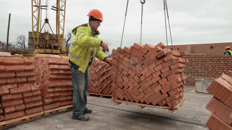 Worker Guiding a Crane Lifting a Pallet of Bricks, a Construction ...