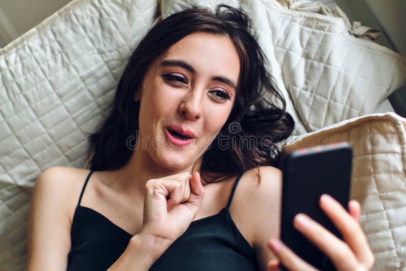 Young Adult Woman Excited Using Mobile Phone While Lying In Bed Stock