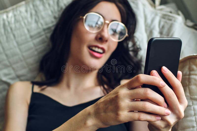 Young Adult Woman Smiling Using Mobile Phone While Lying In Bed Stock