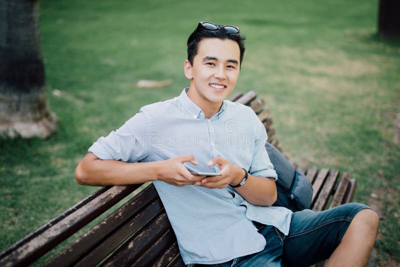 Young Asian Man Sitting On The Bench In The Park With Phone In H Stock