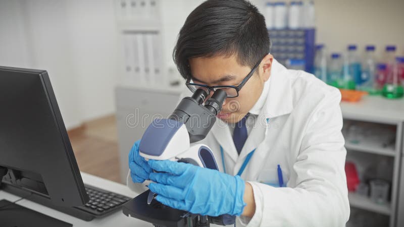 A Young Asian Man in a White Lab Coat Using a Microscope in a ...