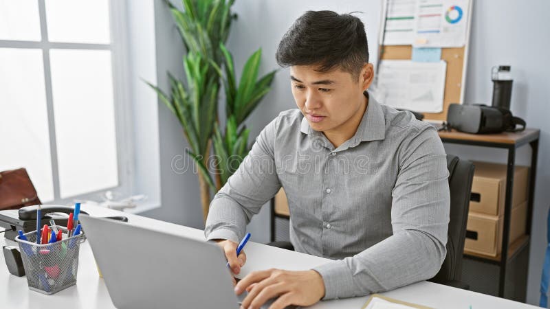A Young Asian Man Working Attentively on His Laptop in a Modern Office ...
