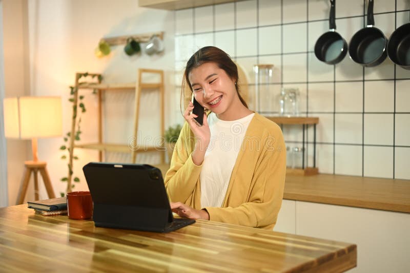 Young Asian Woman Sitting at Kitchen Table Using Laptop and Talking on ...