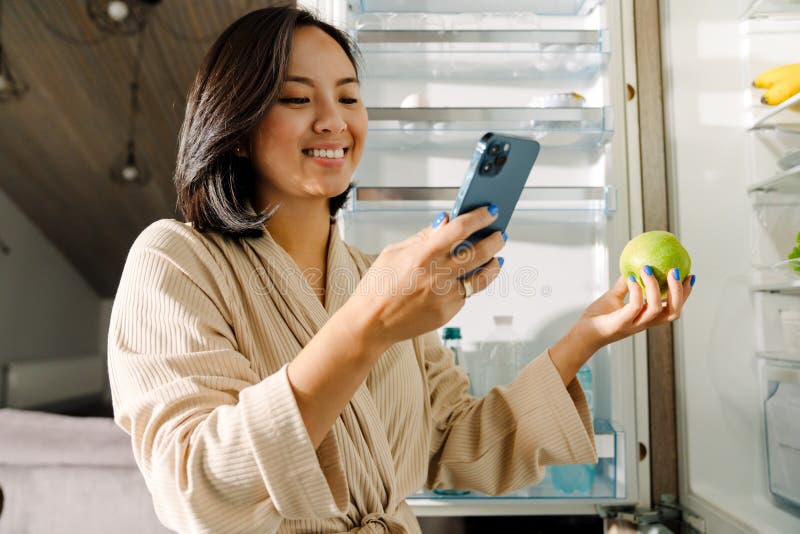 Young Asian Woman Using Mobile Phone while Standing by Open Fridge ...