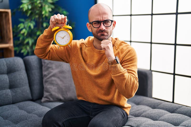 Young Bald Man with Beard Holding Alarm Clock Serious Face Thinking ...
