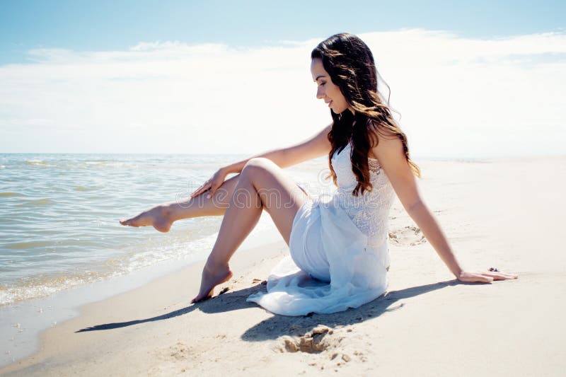 Young Beautiful Brunette Woman In White Dress On The Seashore Sitting