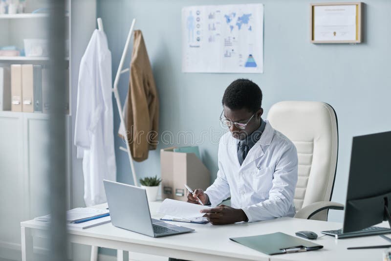 Young Black Doctor Working with Documents at Desk in Office Wearing Lab ...