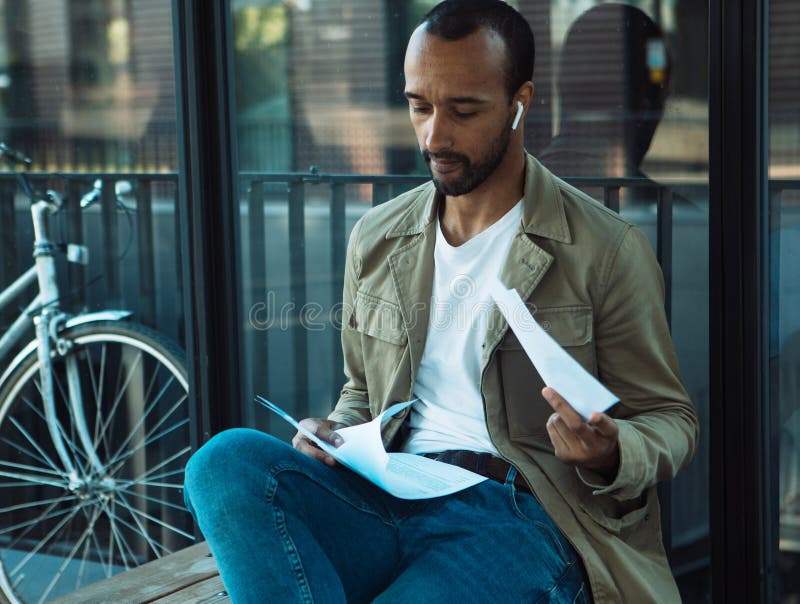 A Young Black Man Looks through Documents while Sitting at a Bus Stop ...