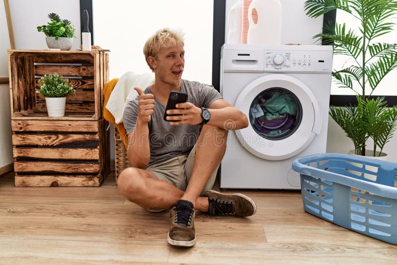 Young Blond Man Doing Laundry Using Smartphone Doing Happy Thumbs Up ...