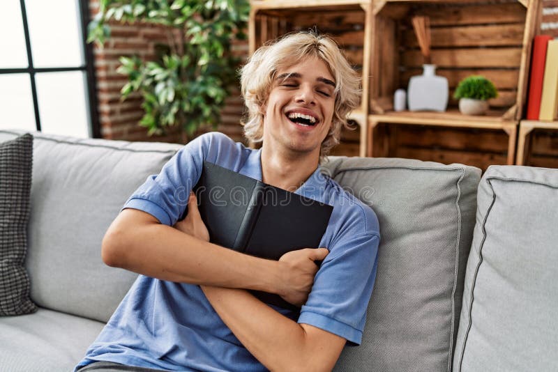 Young Blond Man Hugging Book Sitting on Sofa at Home Stock Photo ...