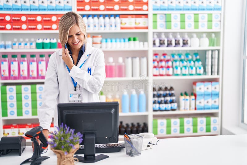 Young Blonde Woman Pharmacist Talking on Smartphone Using Computer at ...