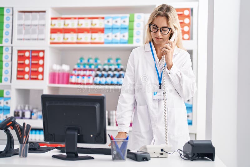 Young Blonde Woman Pharmacist Talking on Telephone Using Computer at ...