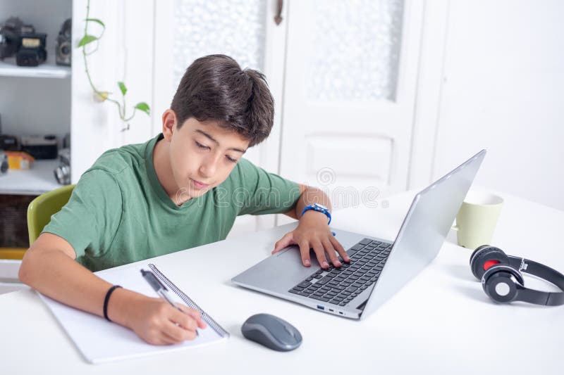A Young Boy is Sitting at a Desk with a Laptop Computer and Writing in ...
