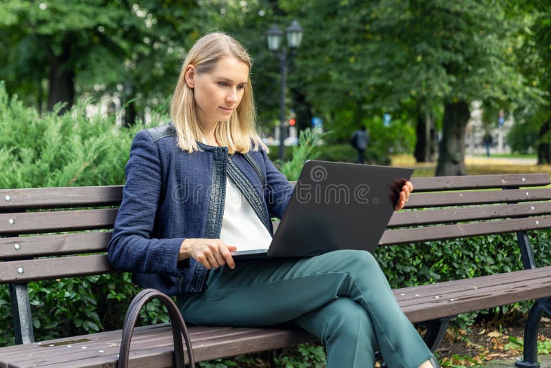 Young Business Woman Sitting On The Bench In The City Park And Using