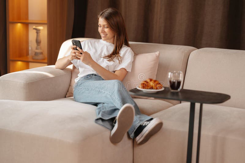 Young Cheerful Woman is Sitting on Sofa and Using Her Phone while Free ...
