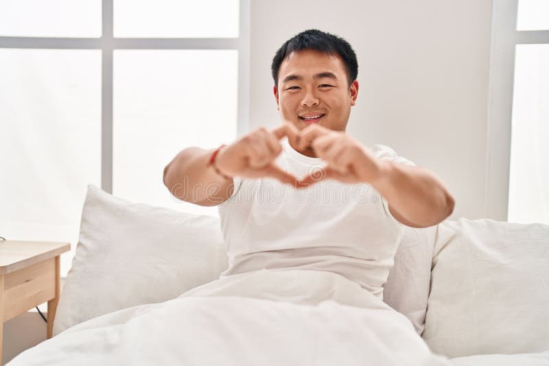 Young Chinese Man Doing Heart Gesture with Hands Sitting on Bed at ...