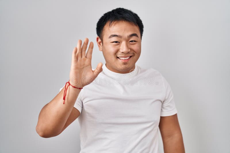 Young Chinese Man Standing Over White Background Waiving Saying Hello ... Young Chinese Man Standing Over White Background Waiving Saying Hello ...