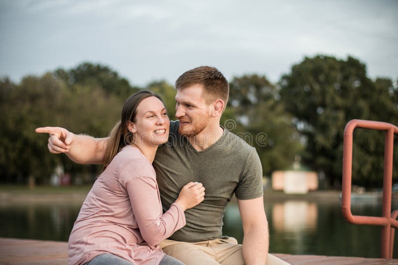 Young Couple In Love Sitting On The Dock Of The Lake Having Fun Stock
