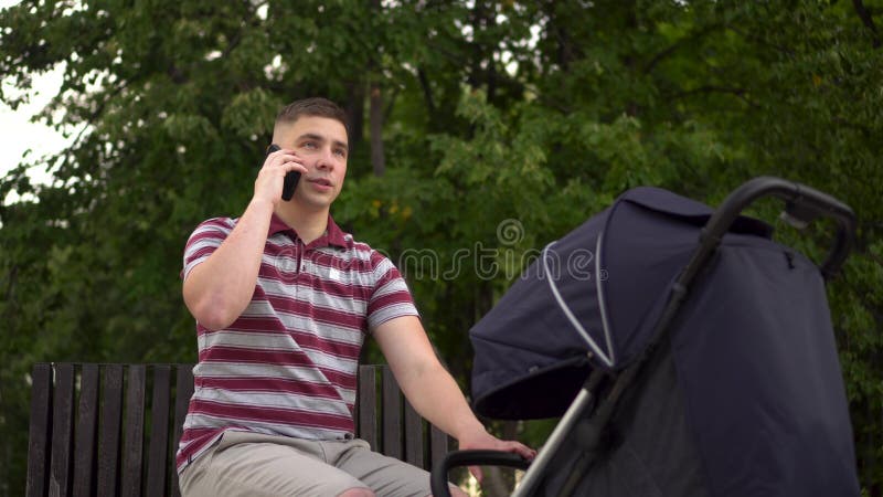 A Young Father is Talking on the Phone and Rocking the Stroller. a Man ...