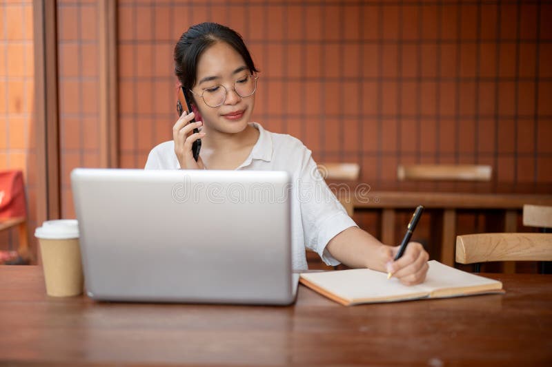 A Focused Asian Woman is Taking Notes in Her Notebook while Talking on ...