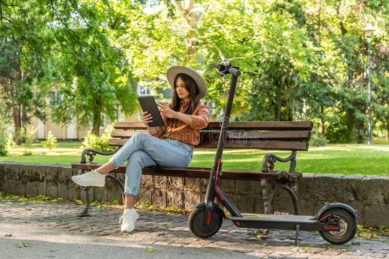 A Young Girl Is Sitting On A Park Bench With The Tablet In Her Hands