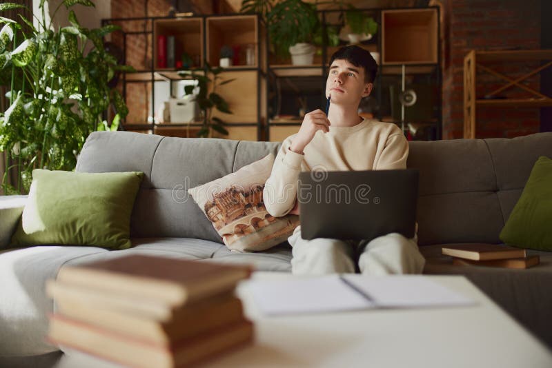 Young Guy Siting on Sofa at Home with Laptop and Doing Homework. Man ...