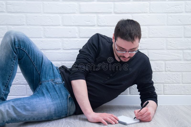 A Young Guy Writes in a Notebook, Lying on a Wooden Floor Against the ...