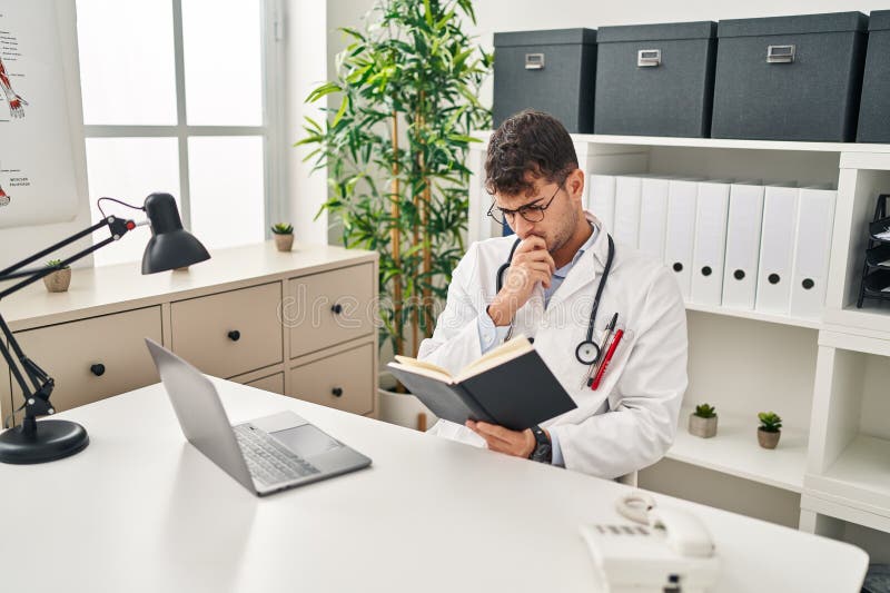 Young Hispanic Man Doctor Using Laptop Reading Book at Clinic Stock ...