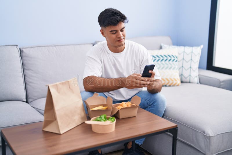 Young Hispanic Man Eating Take Away Food Using Smartphone at Home Stock ...