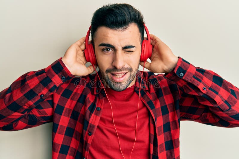 Young Hispanic Man Listening To Music Using Headphones Winking Looking