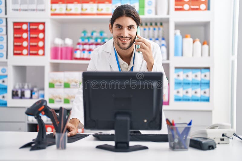 Young Hispanic Man Pharmacist Talking on Smartphone Using Computer at ...