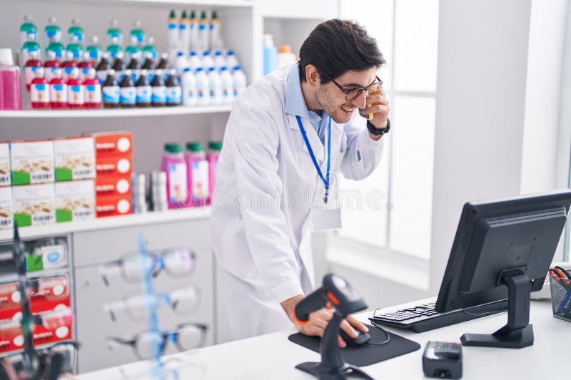 Young Hispanic Man Pharmacist Talking on Smartphone Using Computer at ...