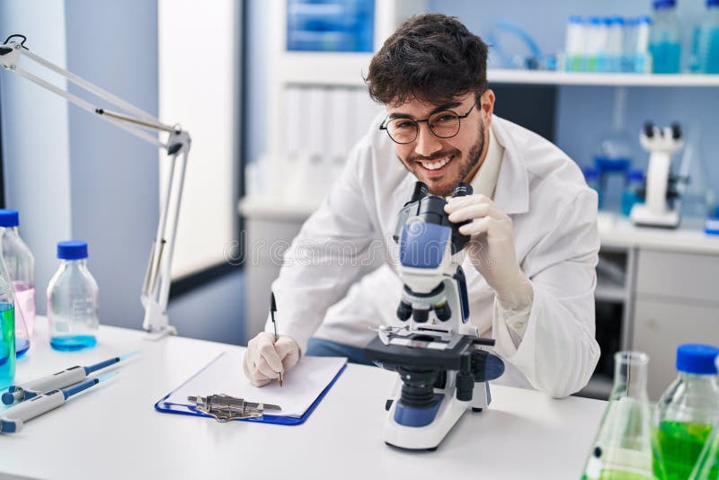 Young Hispanic Man Scientist Writing on Document Using Microscope at ...