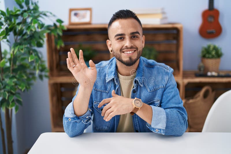 Young Hispanic Man Sitting on Table Saying Hello with Hand at Home ...