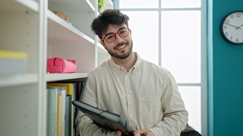 Young Hispanic Man Student Holding Binder Leaning on Shelving at ...