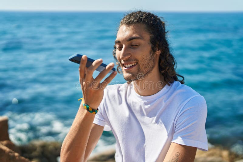 Young Hispanic Man Talking on Smartphone Sitting on Rock at Seaside ...