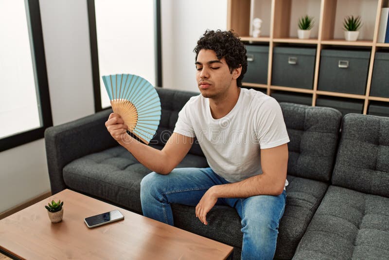 Young Hispanic Man Using Hand Fan Sitting on the Sofa at Home Stock ...