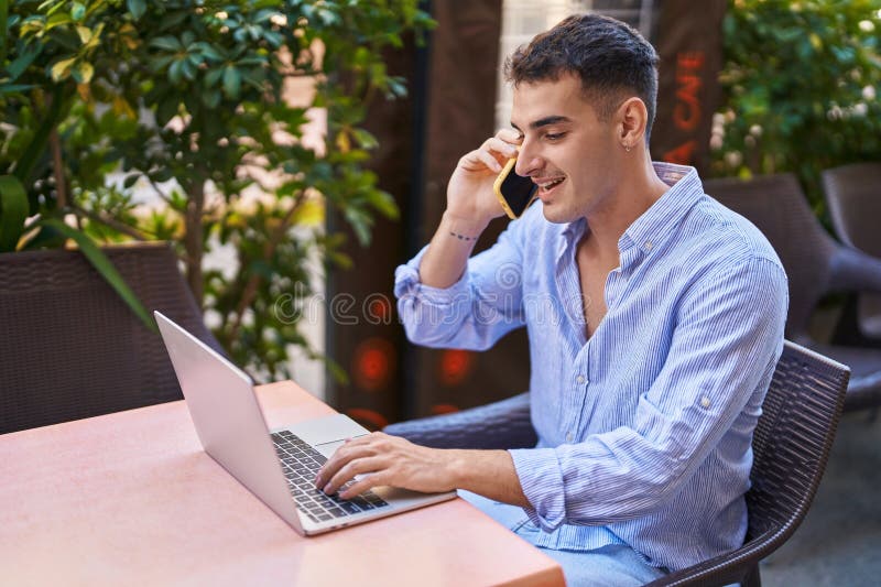 Young Hispanic Man Using Laptop Talking on Smartphone Sitting on Table ...