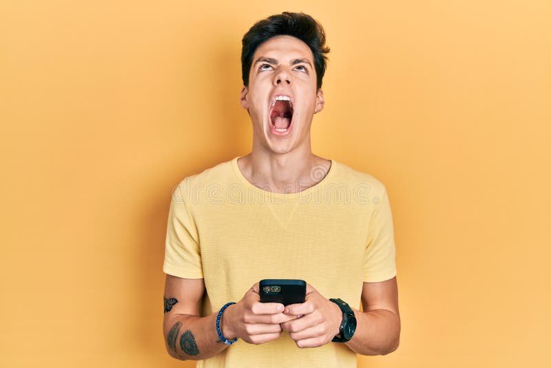 Young Hispanic Man Using Smartphone Typing Message Angry And Mad