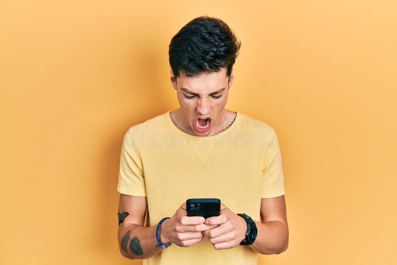 Young Hispanic Man Using Smartphone Typing Message Angry And Mad