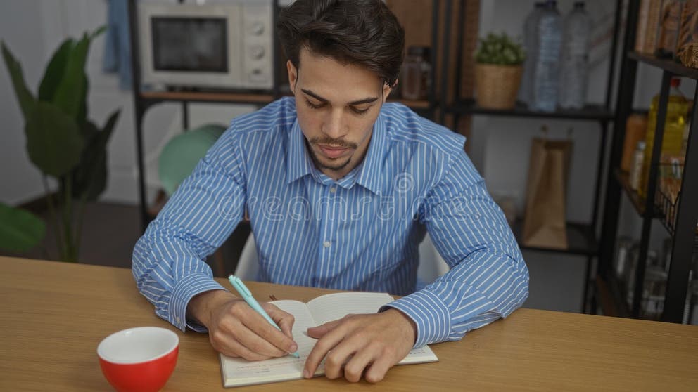 Young Hispanic Man Writing in Notebook at Office Desk with Relaxed ...