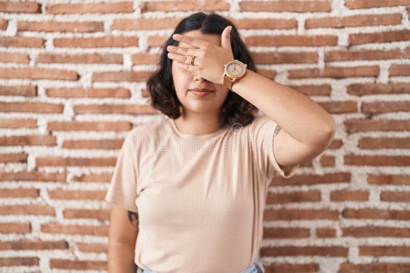 Young Hispanic Woman Standing Over Bricks Wall Covering Eyes With Hand
