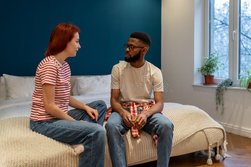 Young Interracial Couple Husband And Wife Sit On Bed At Home Discussing