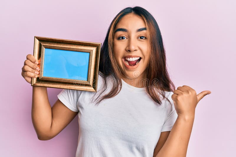 Young Latin Woman Holding Empty Frame Pointing Thumb Up To The Side