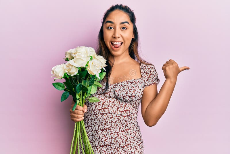 Young Latin Woman Holding Flowers Pointing Thumb Up To The Side Smiling