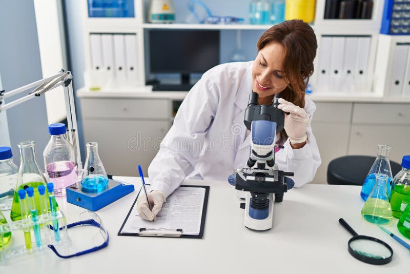 Young Latin Woman Wearing Scientist Uniform Using Microscope Writing on ...