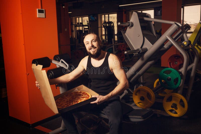 A Young Man with a Beard with a Happy Face Opens a Pizza Box in the Gym ...