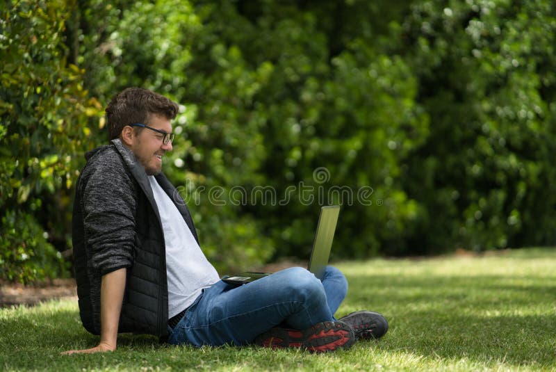 Young Man Looking at His Computer in a Park. he is Sitting on the Green ...