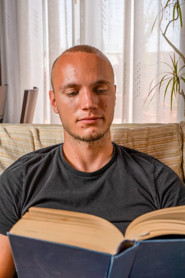 Young Man Reading a Book in His Living Room Sitting on the Couch. Study ...